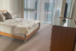 Uma cama bem arrumada em um quarto com uma janela grande, um abajur e uma escrivaninha de madeira.