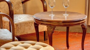 Uma mesa de madeira redonda com dois copos de vinho vazios, uma cadeira estofada com padrão floral e um banquinho com capitonê.