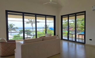 Quarto amplo com grande porta de vidro deslizante que dá para uma varanda com vista para o oceano e palmeiras.