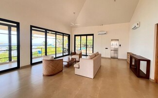 Uma sala de estar ampla, com grandes janelas, um sofá, uma mesa de centro e uma poltrona de vime.