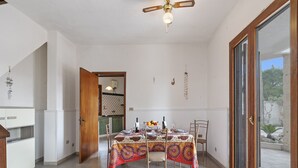 Uma área de refeições com uma mesa posta para dois, um ventilador de teto e vista através de uma porta de vidro.