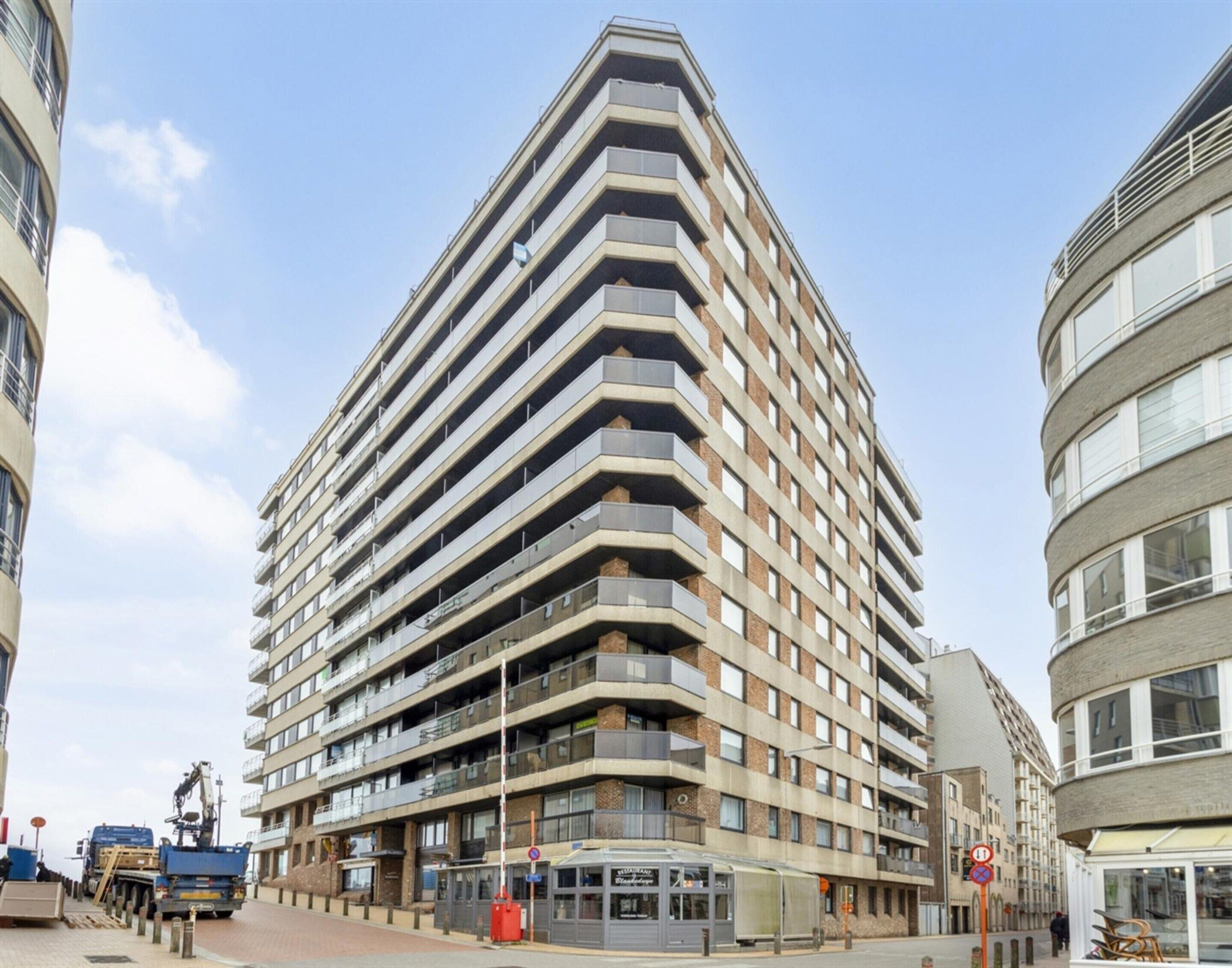 Um edifício residencial com vários andares, varandas, um ponto de ônibus e um veículo de construção por perto.