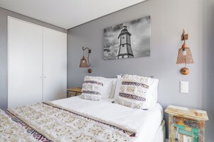 Quarto com cama, pintura de um farol, luminária pendente e uma pequena mesa.