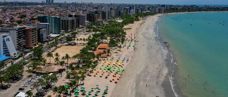 Paisagem costeira urbana com uma praia repleta de guarda-sóis e palmeiras.