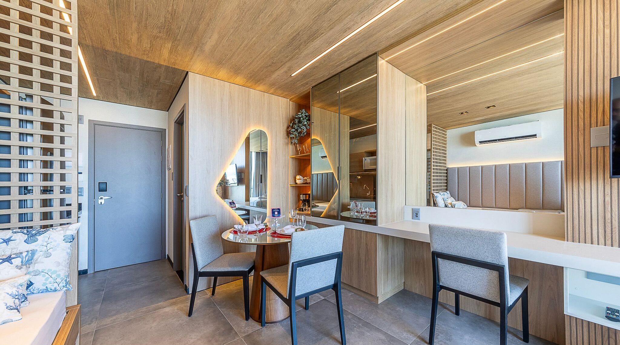 Quarto moderno e bem iluminado, com teto de madeira, área de jantar com duas cadeiras e uma mesa, um espelho grande e um ar condicionado embutido.