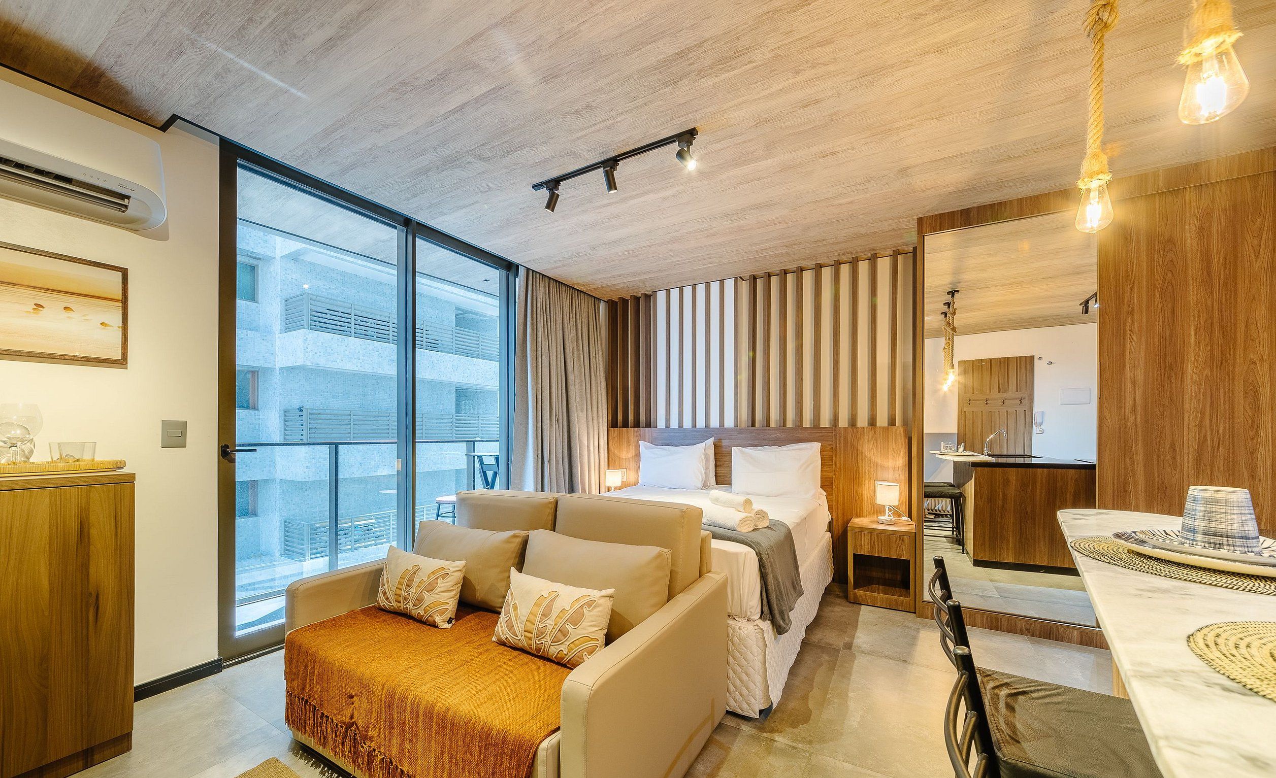 Quarto de hotel moderno com uma cama grande, área de estar com sofá e poltronas, pequena escrivaninha e vista da cidade através de grandes janelas.