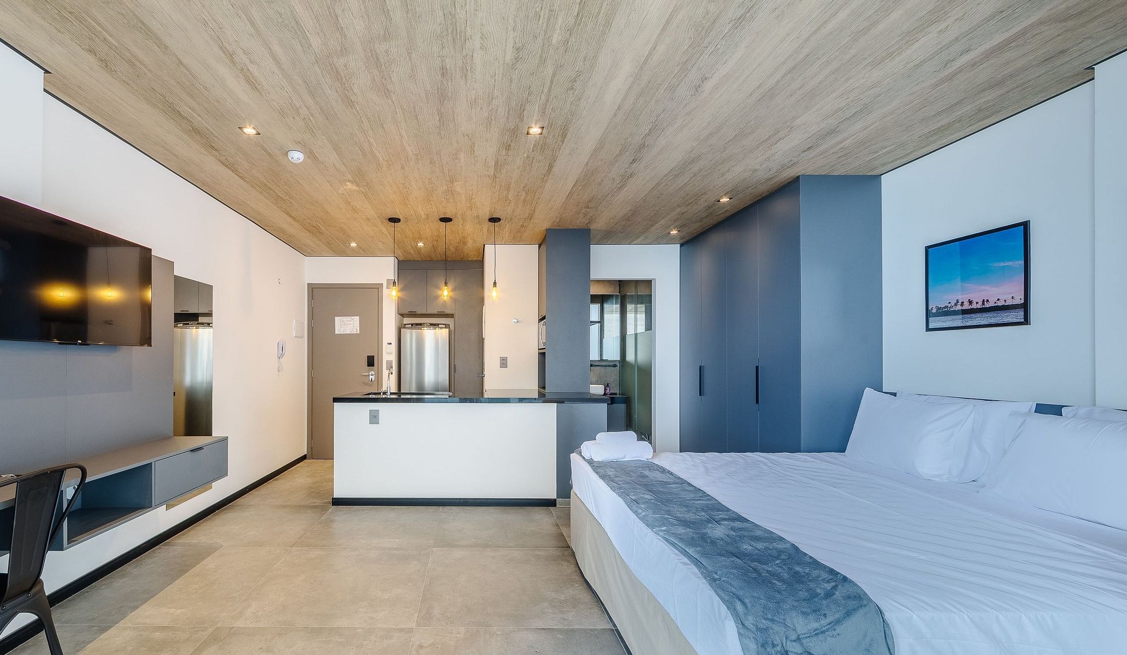Quarto de hotel moderno com teto de madeira, cama, televisão e cozinha compacta.