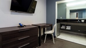 Quarto de hotel com penteadeira de madeira escura, uma cadeira branca, televisão de tela plana na parede e um espelho grande sobre a pia.