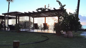 Uma área de estar coberta ao ar livre, com cadeiras e mesas, rodeada por vegetação e vista para o mar.