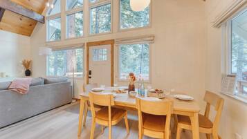 Uma área de jantar moderna com mesa e cadeiras de madeira, janelas amplas e um sofá cinzento.