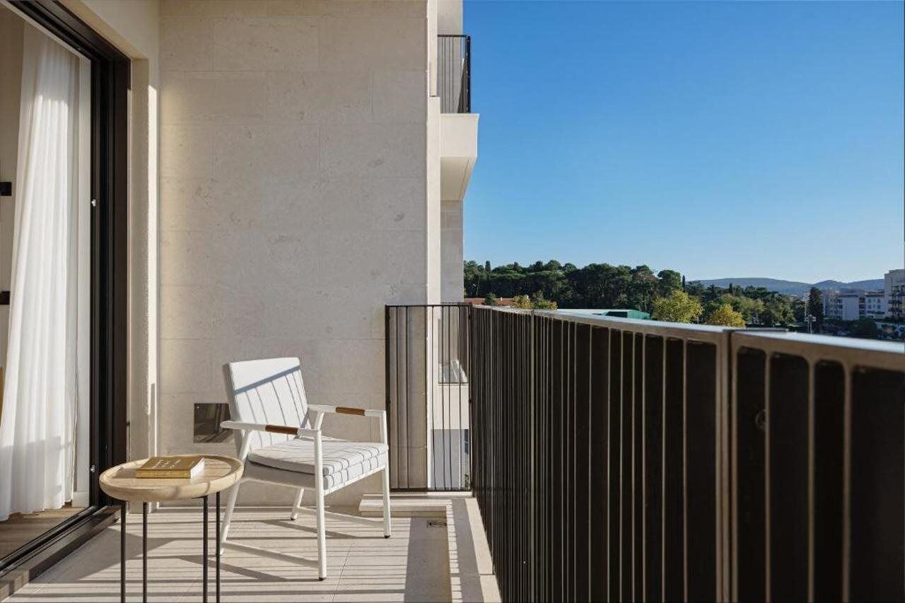Uma varanda com uma cadeira branca e uma mesa pequena, guarda-corpo de metal e vista para a paisagem urbana com áreas verdes e edifícios.