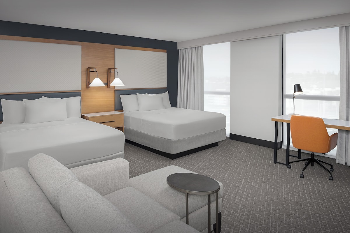 Quarto de hotel moderno com duas camas, uma escrivaninha, uma cadeira laranja e uma pequena mesa redonda.