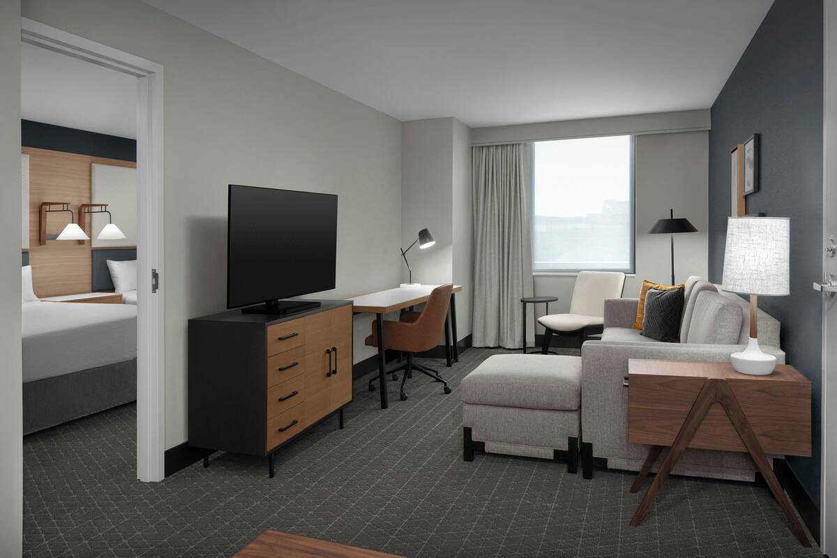 Quarto de hotel moderno com cama, escrivaninha, televisão, sofá e janela com cortinas.