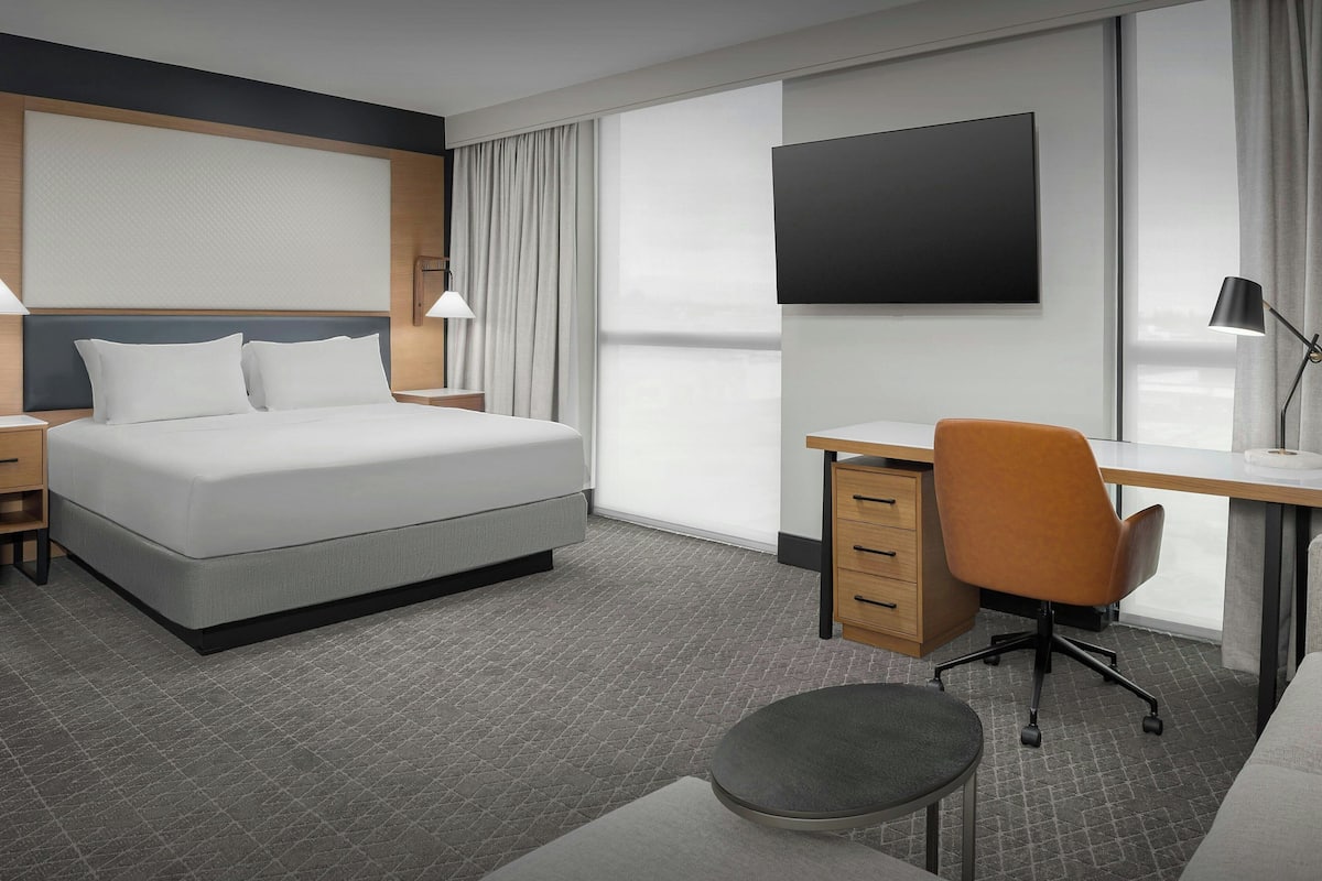 Quarto de hotel moderno com cama, escrivaninha, cadeira e grandes janelas.