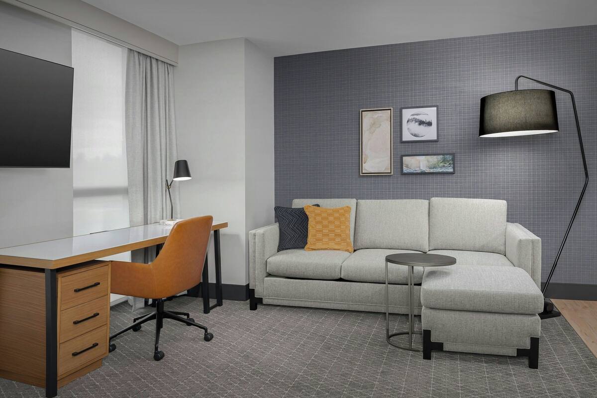 Quarto de hotel moderno com uma mesa, um sofá, uma pequena mesa redonda e um abajur.