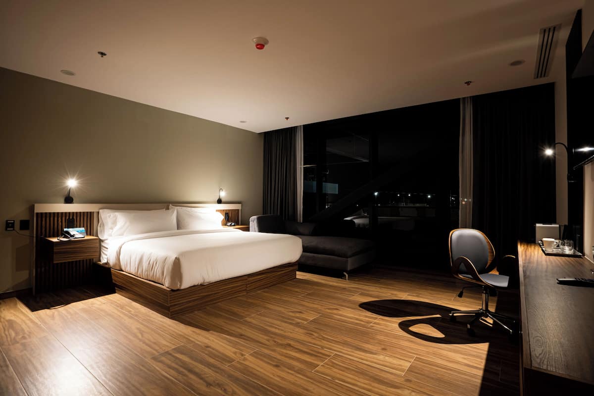 Quarto de hotel moderno com uma cama grande, escrivaninha, cadeira e vista da cidade à noite.