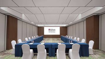 Uma sala de conferência com uma mesa comprida, cadeiras brancas e o logotipo da Hilton Garden Inn na tela.