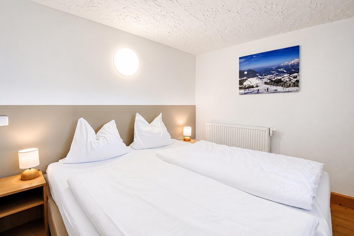 Quarto de hotel com duas camas, uma luminária redonda na parede, um radiador e um quadro emoldurado.