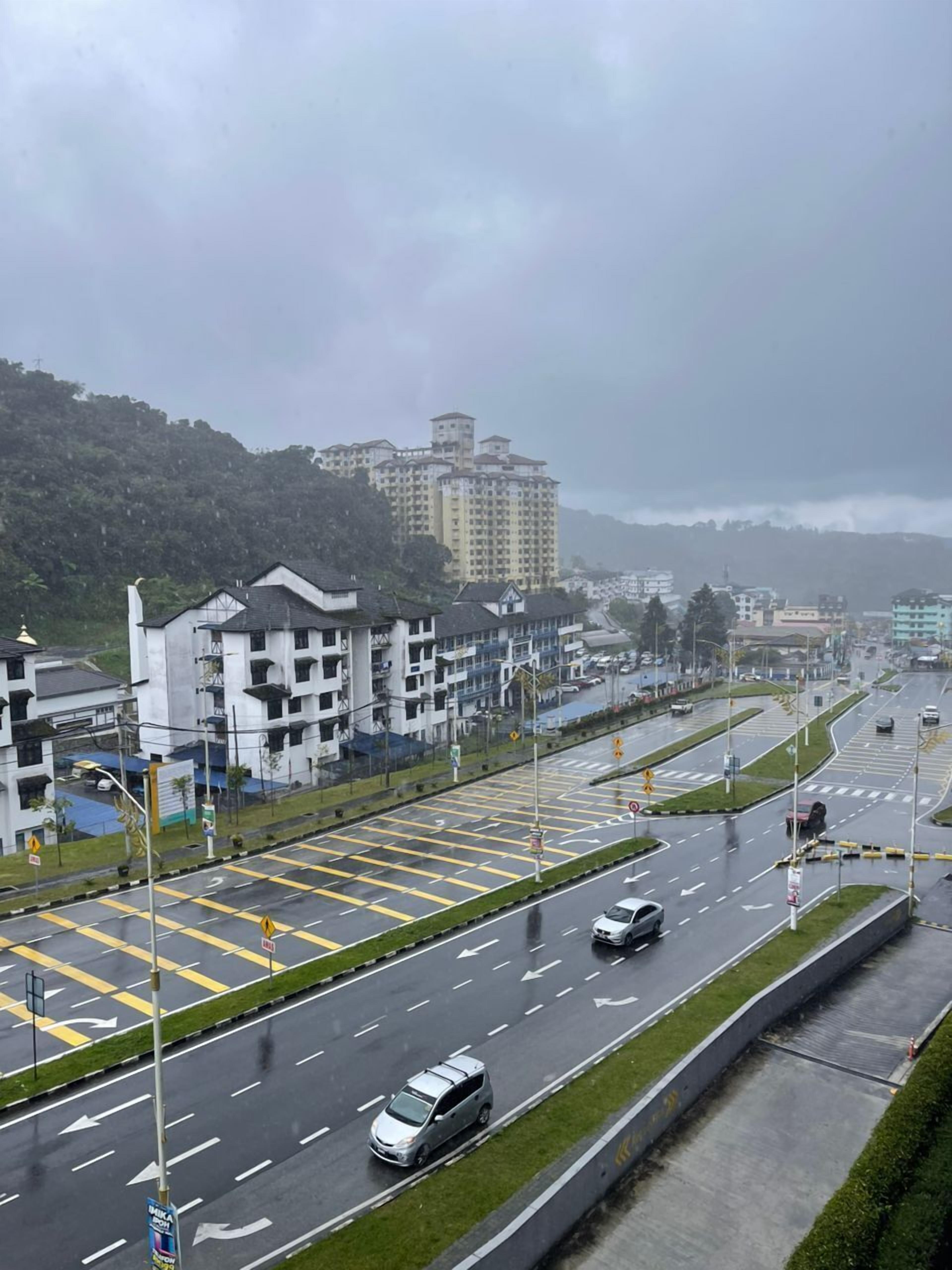 Uma estrada com várias faixas, carros, uma faixa de pedestres e edifícios ao fundo.