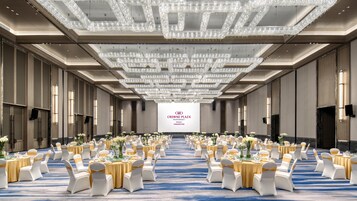 Uma sala de conferências ampla e bem iluminada, com fileiras de mesas redondas, cada uma com cadeiras brancas e fitas douradas, e uma tela grande na frente exibindo o logotipo Crowne Plaza RUGAO.