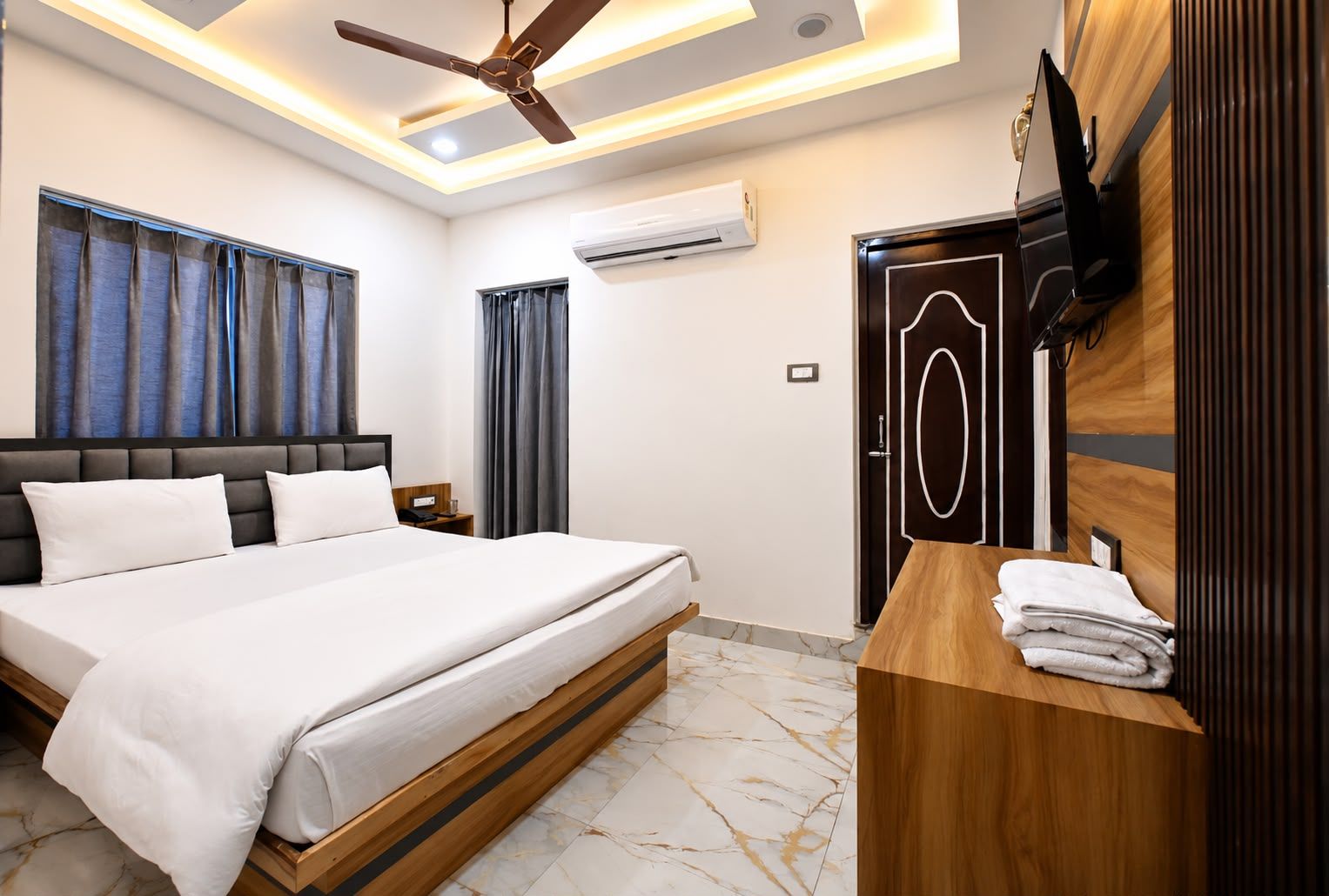 Quarto de hotel com uma cama, mesa de cabeceira de madeira com toalhas, ventoinha no teto e ar condicionado.