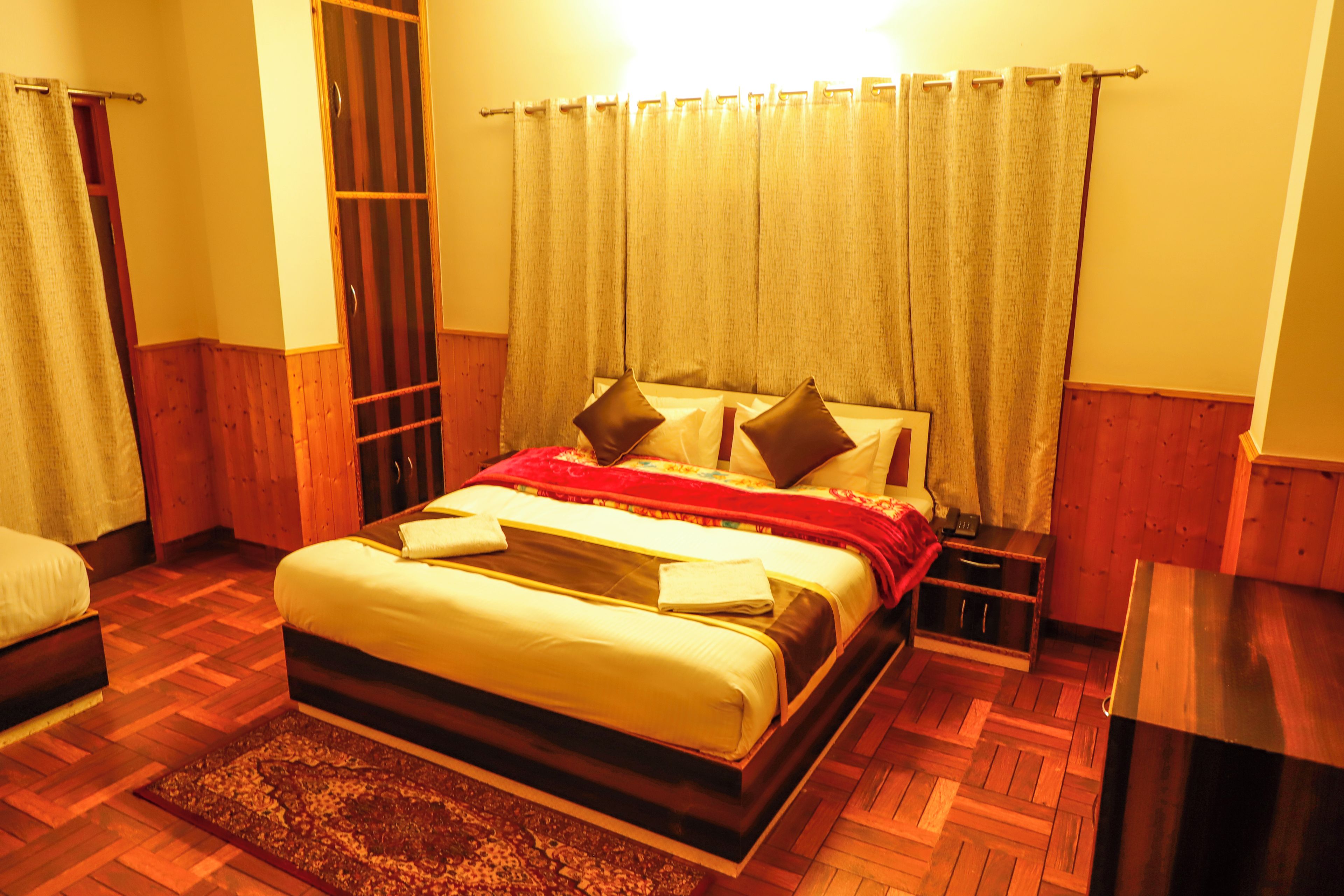 Quarto de hotel com uma cama grande, mesa de cabeceira, piso de madeira e janela com cortinas.