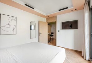 Quarto de hotel com cama, televisão fixada na parede, porta para uma área de refeições e uma grande pintura abstrata.