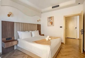 Quarto de hotel com uma cama grande, cabeceira de madeira, mesa de cabeceira e luminária fixada na parede.