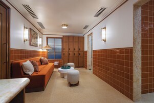 Um corredor com parede revestida de azulejos marrons, um sofá marrom com almofadas, uma mesinha redonda pequena e um banquinho branco.
