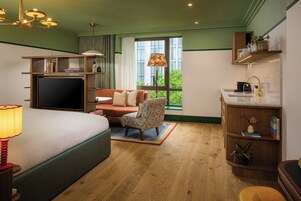 Quarto de hotel com cama, sofá, poltrona, televisão, escrivaninha, luminária, janela com vista e pia.