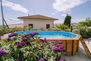 Uma banheira de madeira com piscina azul cristalina, rodeada por vegetação exuberante e flores roxas, com uma casa ao fundo.