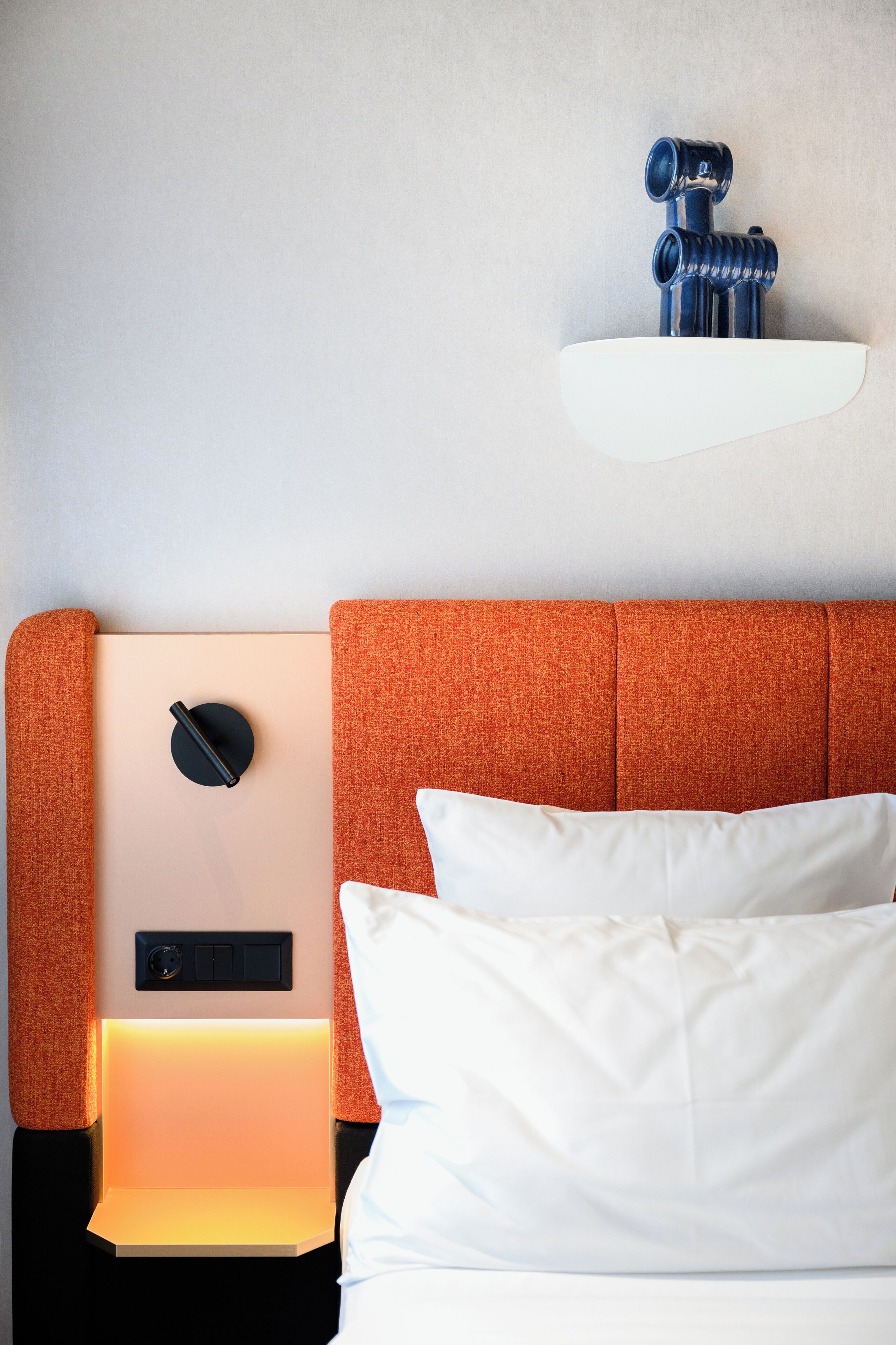 Quarto de hotel moderno com uma cama, mesa de cabeceira, luminária de parede e prateleira com objetos decorativos.