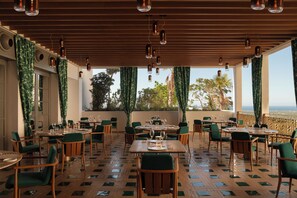 Um restaurante com mesas e cadeiras de madeira, grandes janelas com cortinas e luminárias pendentes.