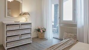 Quarto com cômoda branca, espelho, abajur, cesta de vime com flores, cama com cobertor e vista para o exterior pela janela.