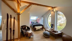 Um amplo quarto no estilo loft, com claraboia, vigas de madeira, cama, poltrona, banco e esquis encostados na parede.