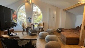 Uma sala de estar moderna com televisão de tela plana, pufes redondos, um sofá e um espaço de jantar com cadeiras e uma mesa.