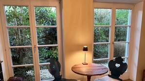 Um quarto com piso de madeira, uma mesa redonda com abajur, duas cadeiras e um banquinho pequeno.