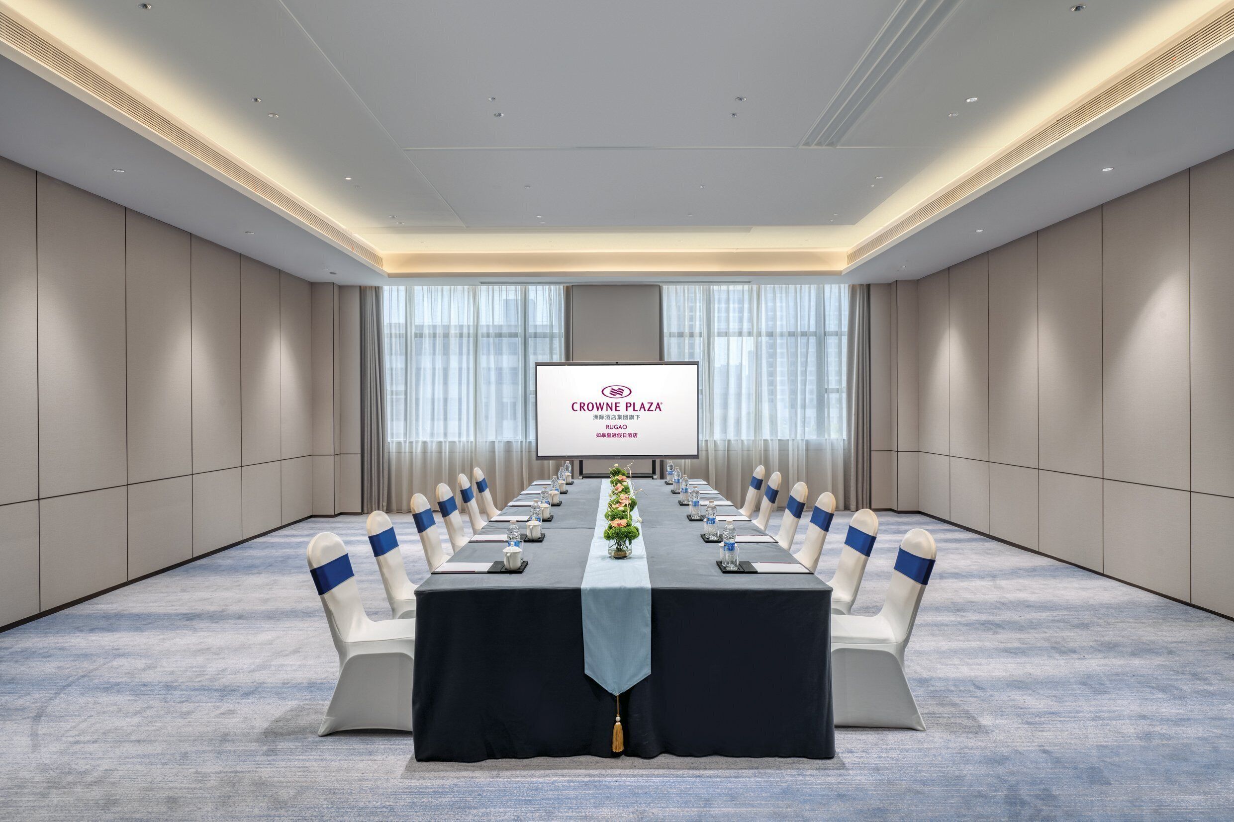 Uma sala de conferências com uma mesa comprida, cadeiras e uma tela exibindo "CROWNE PLAZA".