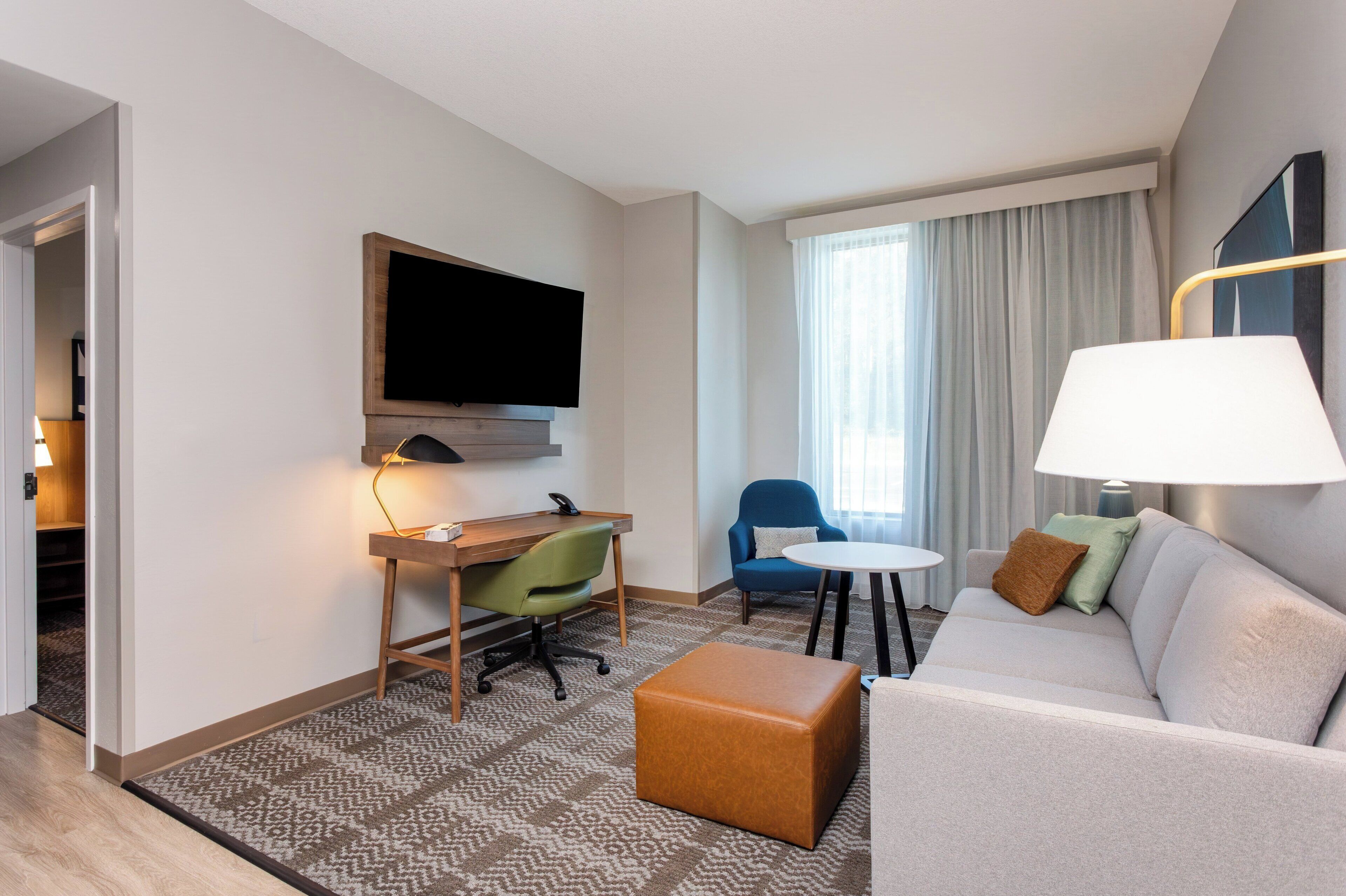 Quarto de hotel com sofá, escrivaninha com cadeira, televisão, luminária e janela com cortinas.