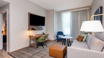 Quarto de hotel com sofá, escrivaninha com cadeira, televisão, luminária e janela com cortinas.