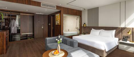 Quarto de hotel moderno com cama grande, sofá, pequena mesa com frutas, closet espaçoso e banheiro com banheira e chuveiro.