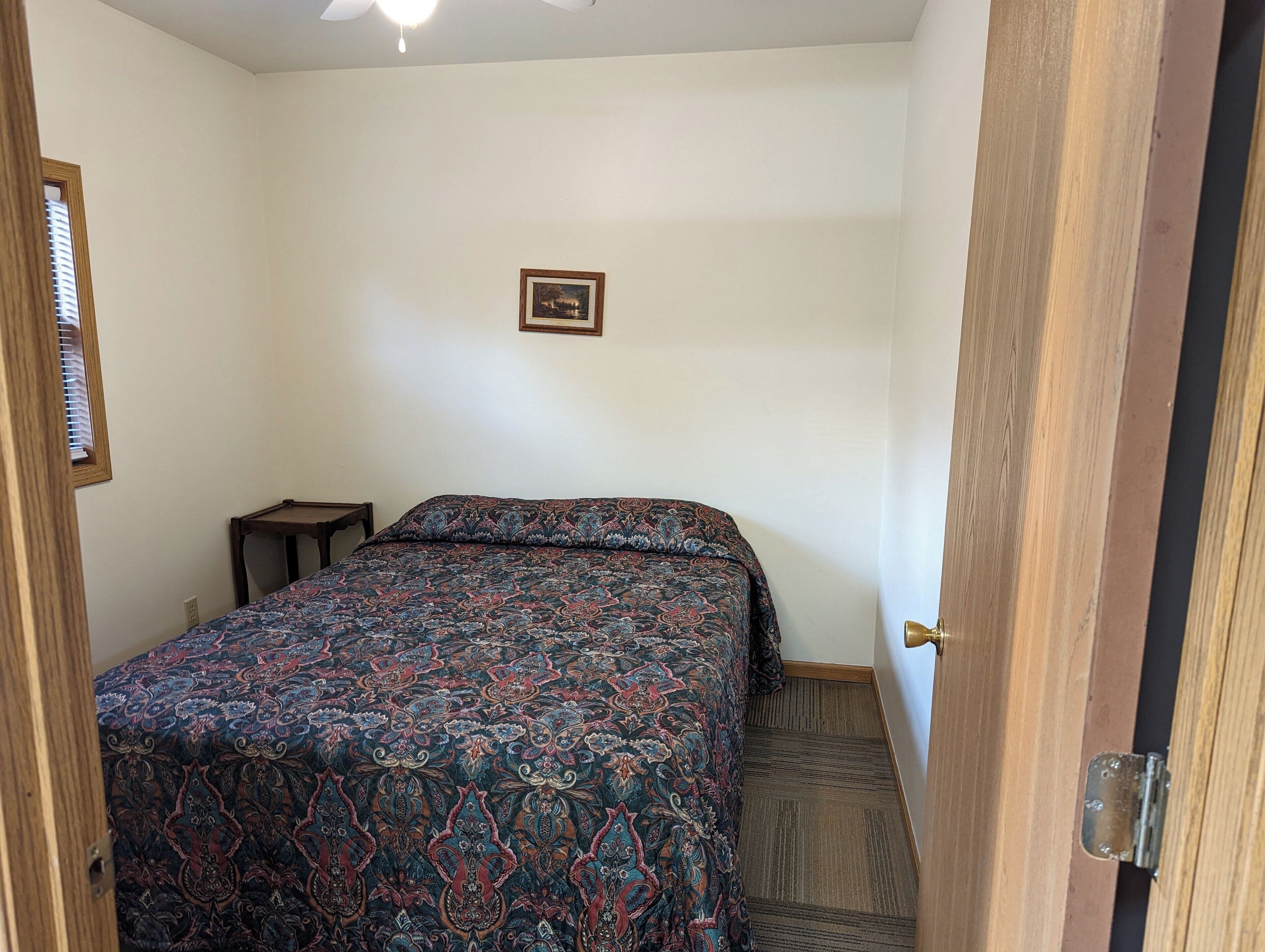 Quarto pequeno com uma cama de solteiro, colcha estampada, uma pequena mesa de cabeceira, um quadro na parede e uma porta com puxador de latão.