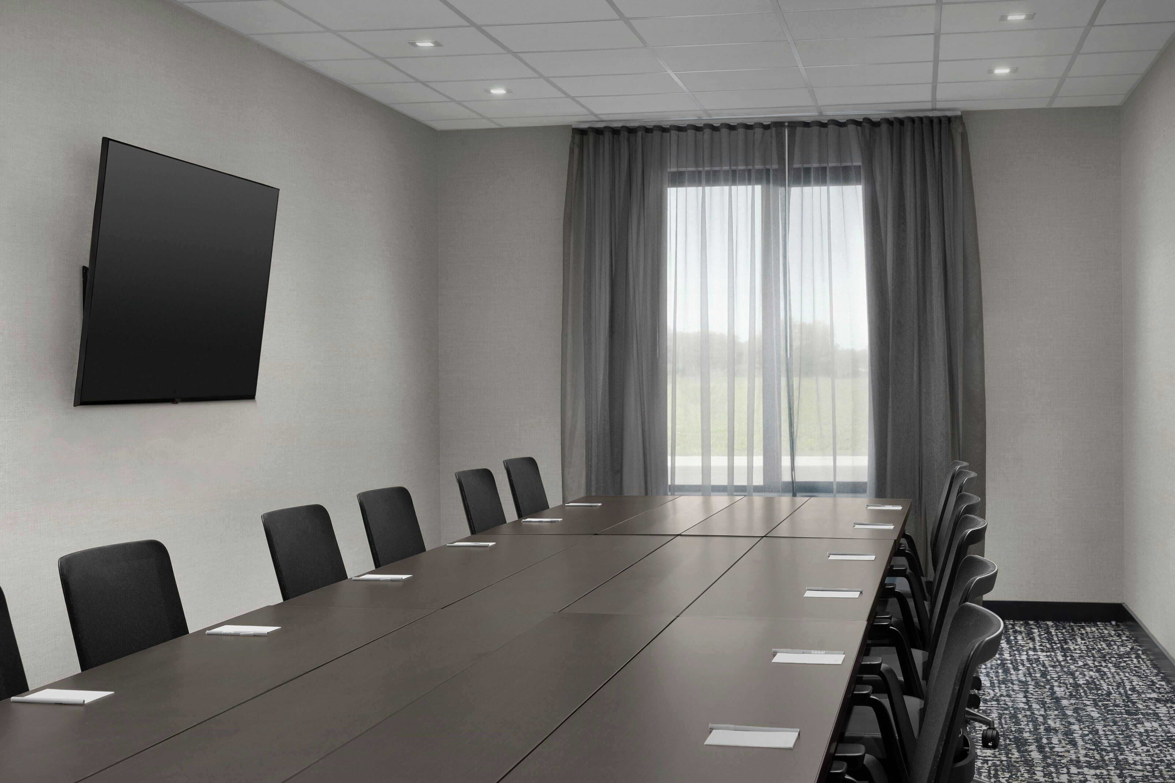 Uma sala de conferências com uma mesa comprida, cadeiras pretas, uma TV de tela plana na parede e uma grande janela com cortinas.
