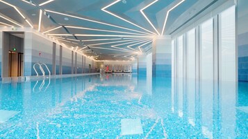 Uma piscina interior moderna com iluminação de teto geométrica e paredes de vidro transparente.