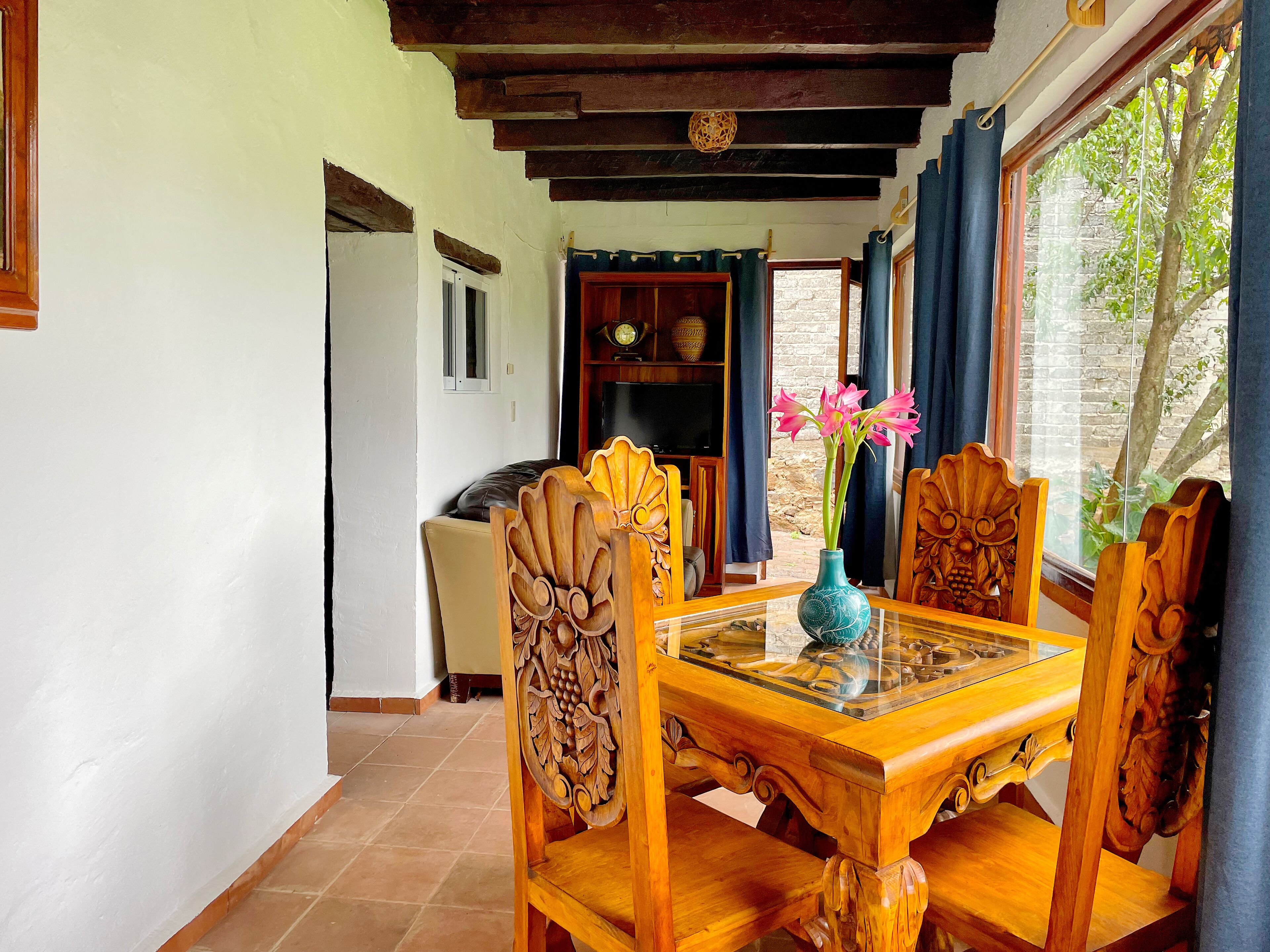 Uma área de refeições com uma mesa e cadeiras de madeira, um vaso com flores e vista para o exterior através de uma janela.