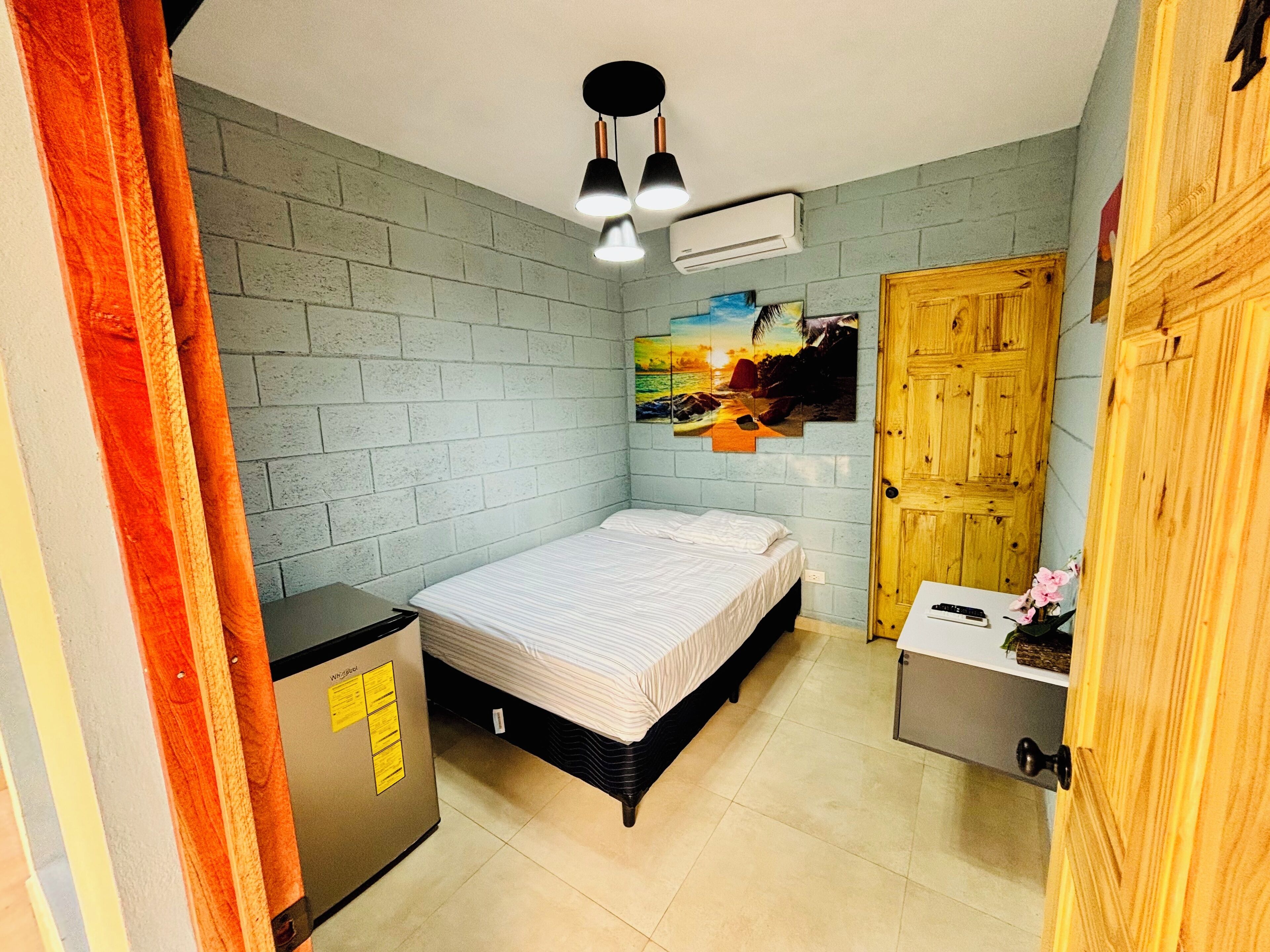Quarto compacto com cama, guarda-roupa de madeira, pequena escrivaninha e ar-condicionado fixado na parede.
