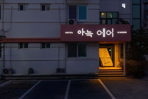 Entrada de hotel com placa em coreano, aparelhos de ar condicionado e sinalização de estacionamento.