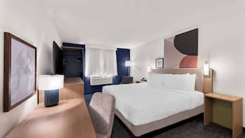 Quarto de hotel moderno com uma cama grande, uma escrivaninha, uma cadeira, uma mesinha de cabeceira e uma luminária.