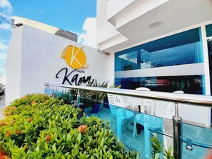 Exterior de hotel com logotipo em destaque, área de estar ao ar livre e guarda-corpo de vidro.