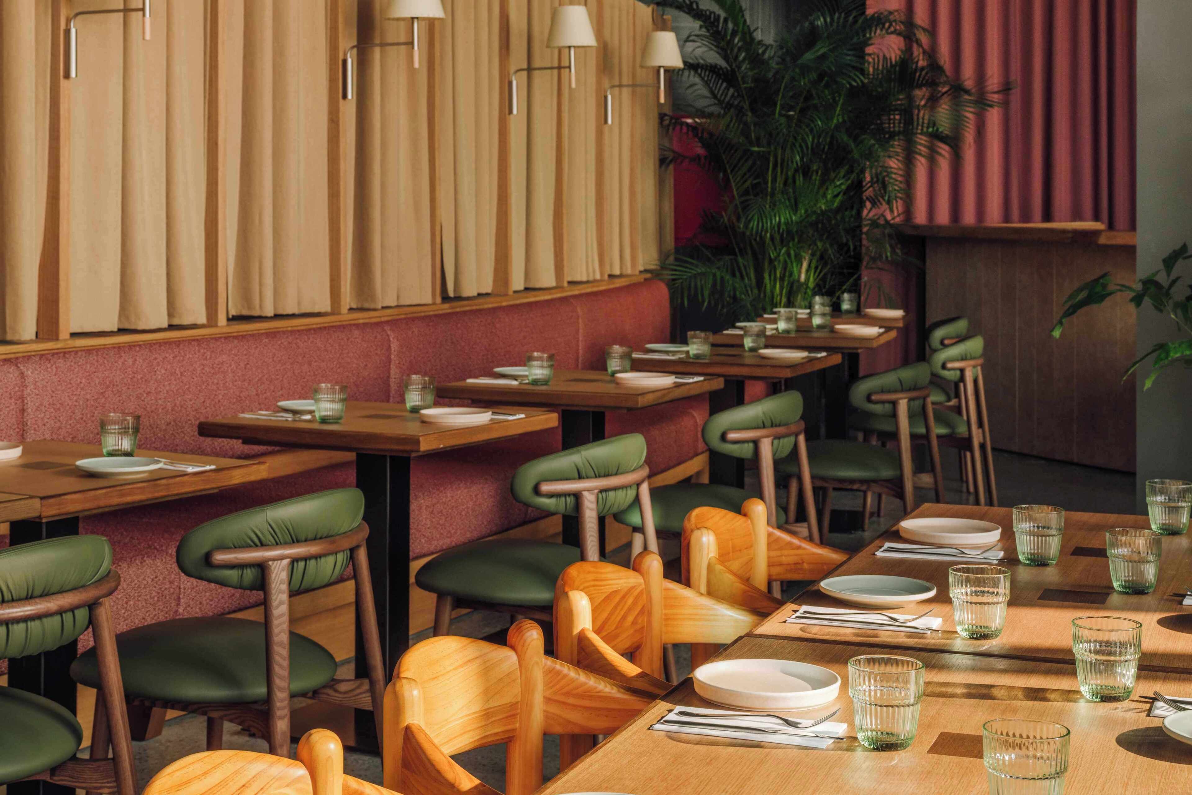Interior de restaurante com mesas e cadeiras de madeira, assentos acolchoados verdes e um fundo de cortina vermelha.